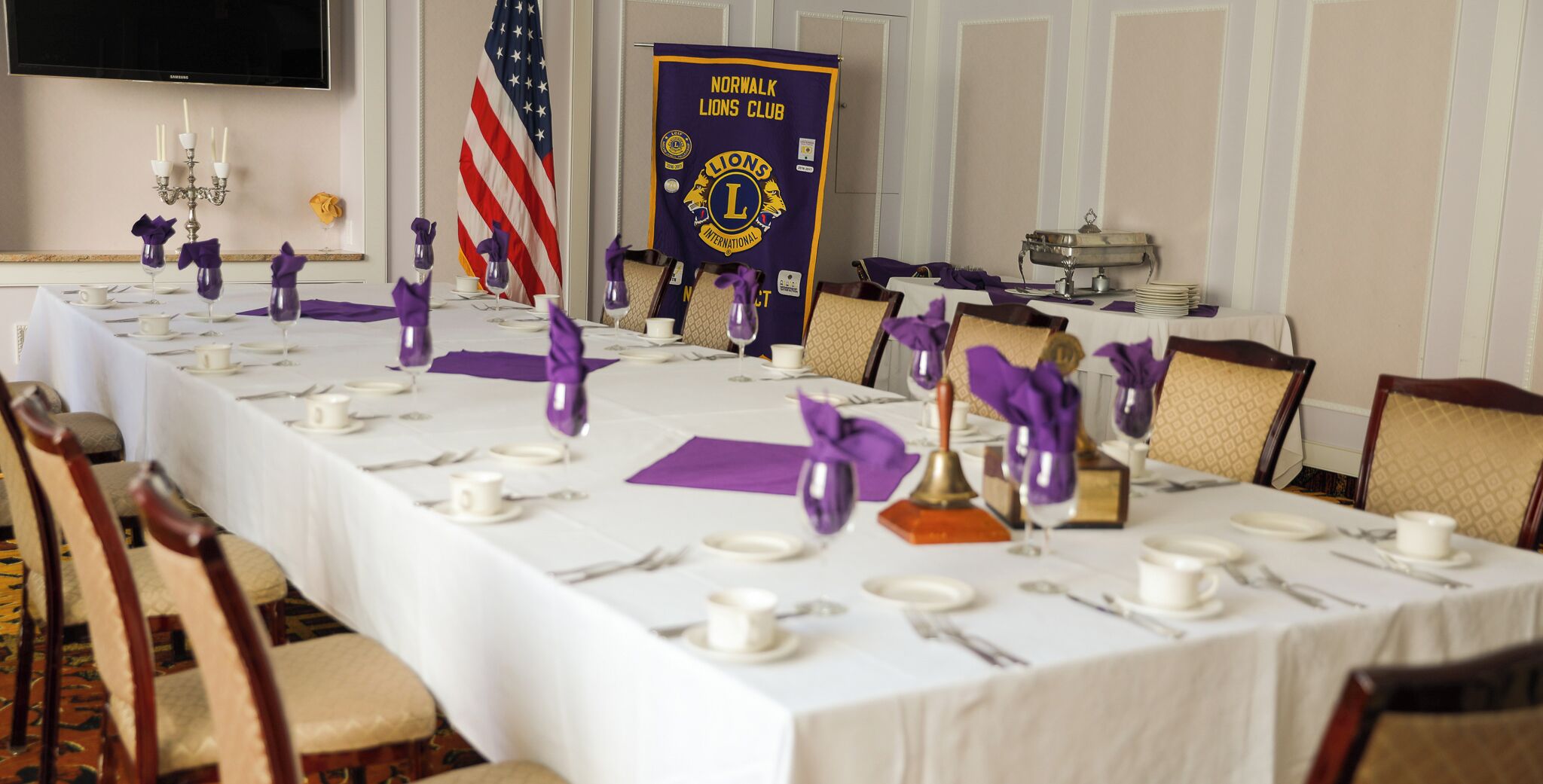 Uma sala de conferências com uma mesa comprida preparada para uma reunião, decorada com guardanapos roxos e louça branca. Uma bandeira americana e um banner do Norwalk Lions Club são visíveis ao fundo.