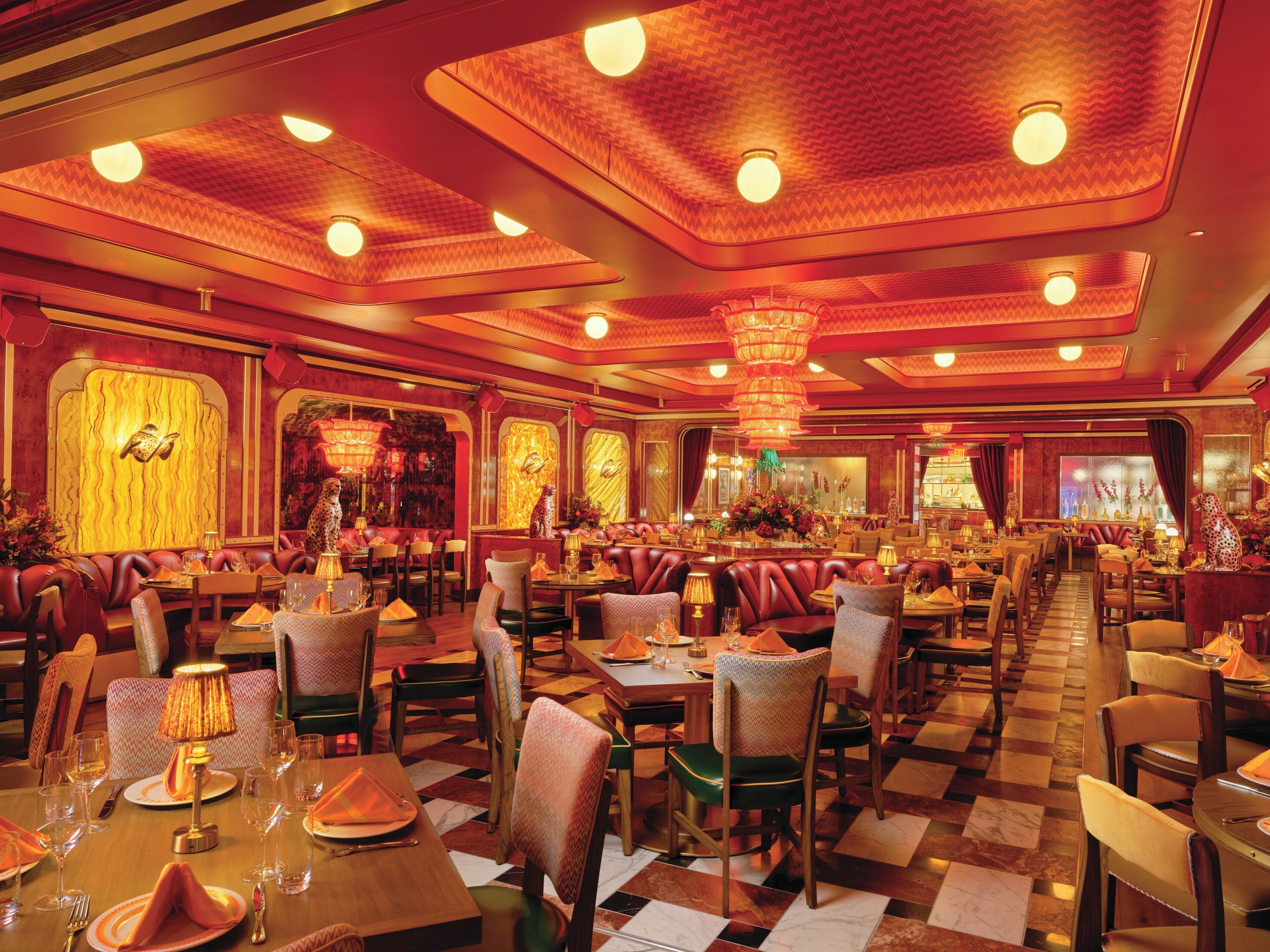 Restaurante com decoração em vermelho e dourado, assentos de couro e lustres. Mesas dispostas com copos de vinho e pratos.