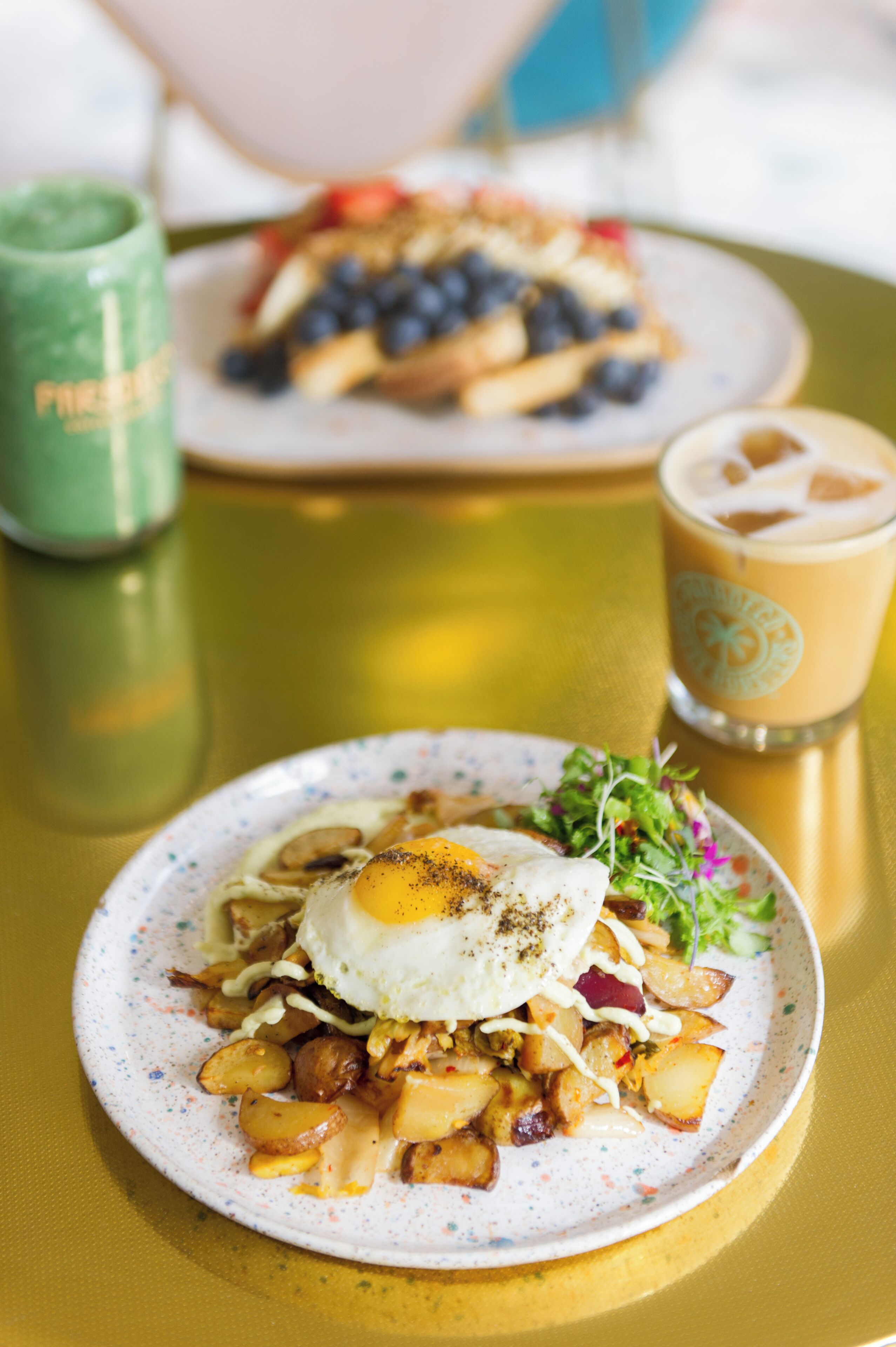 Prato de café da manhã com ovos, batatas e vegetais, um copo de café gelado e um smoothie verde sobre uma mesa amarela.