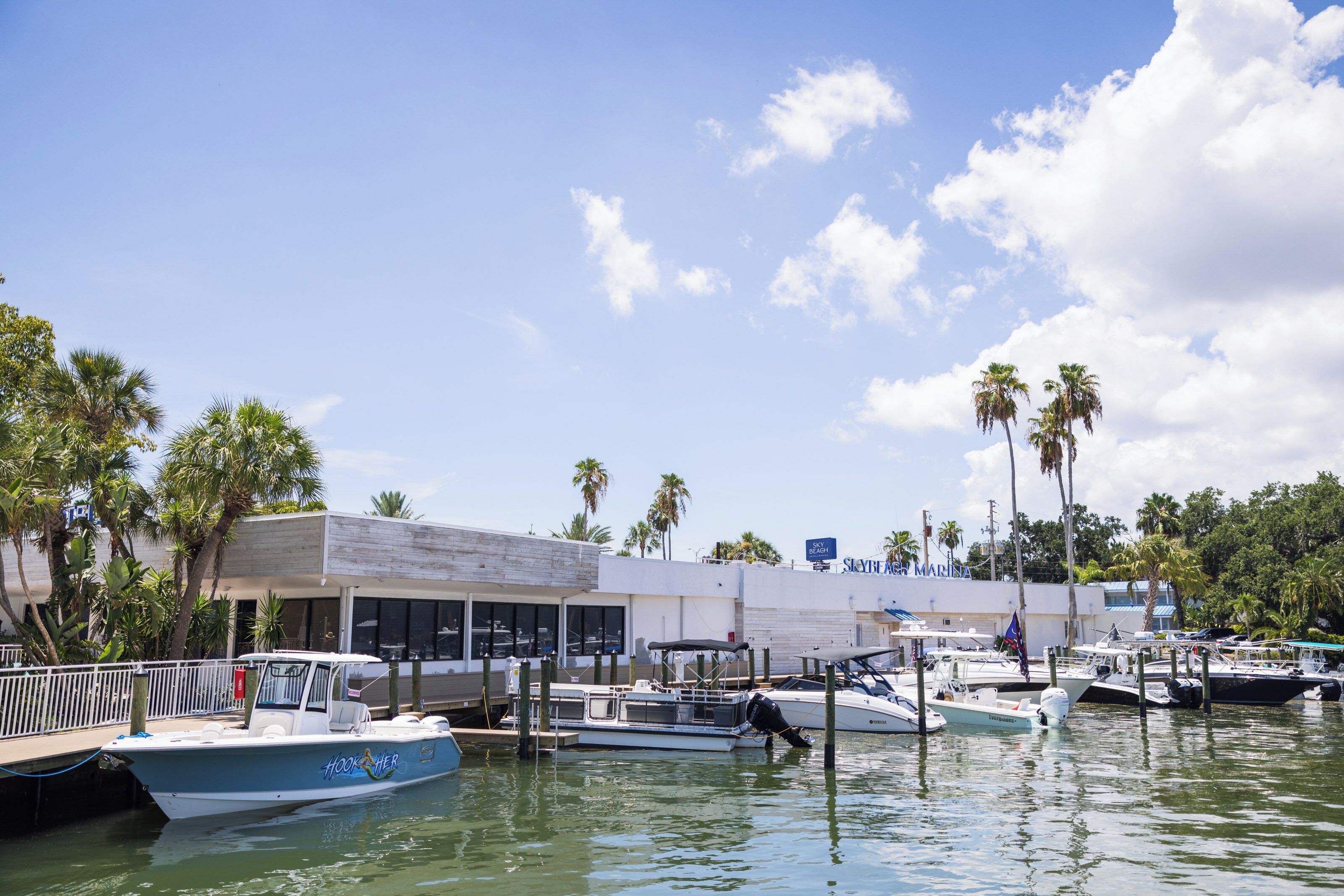 Uma marina com vários barcos atracados, um edifício com a placa 'Skyline Marina' e palmeiras ao fundo.