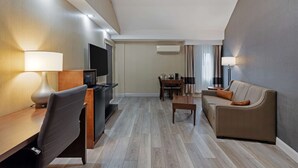 Quarto de hotel com televisão de tela plana, uma mesa com abajur, um sofá, uma mesa de jantar com cadeiras e um aparelho de ar condicionado.