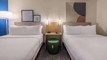 Quarto de hotel com duas camas, um banquinho verde, uma mesa de cabeceira com abajur e obras de arte nas paredes.