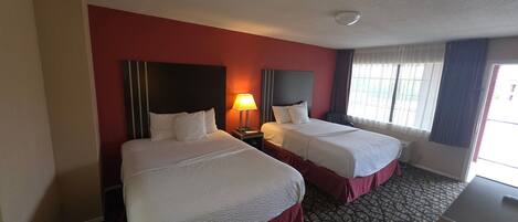 Quarto de hotel com duas camas, uma mesa de cabeceira com abajur, janela com cortinas e radiador.