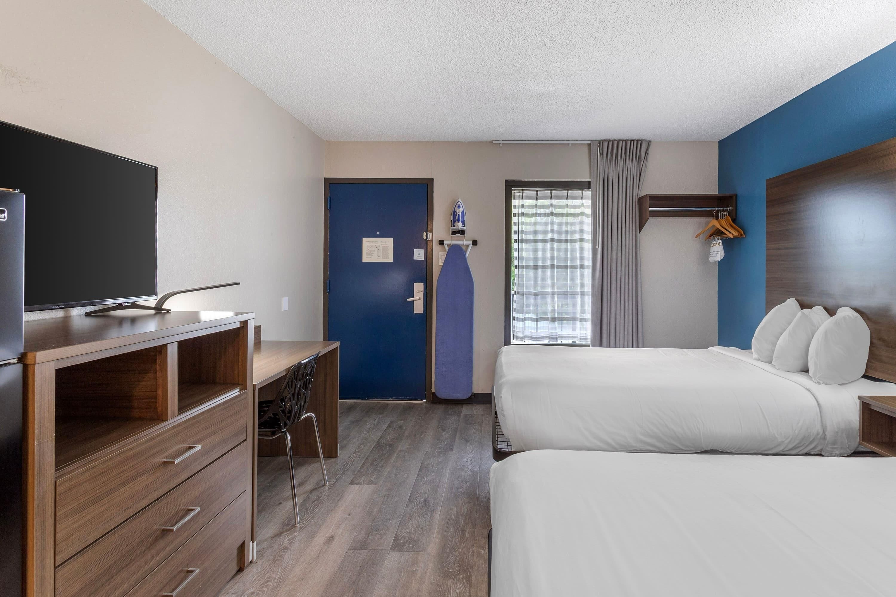 Quarto de hotel com duas camas, uma escrivaninha com gavetas, televisão de tela plana, uma porta azul e uma janela com cortinas.