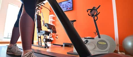 Uma academia com equipamentos de exercícios, esteira e televisão na parede.