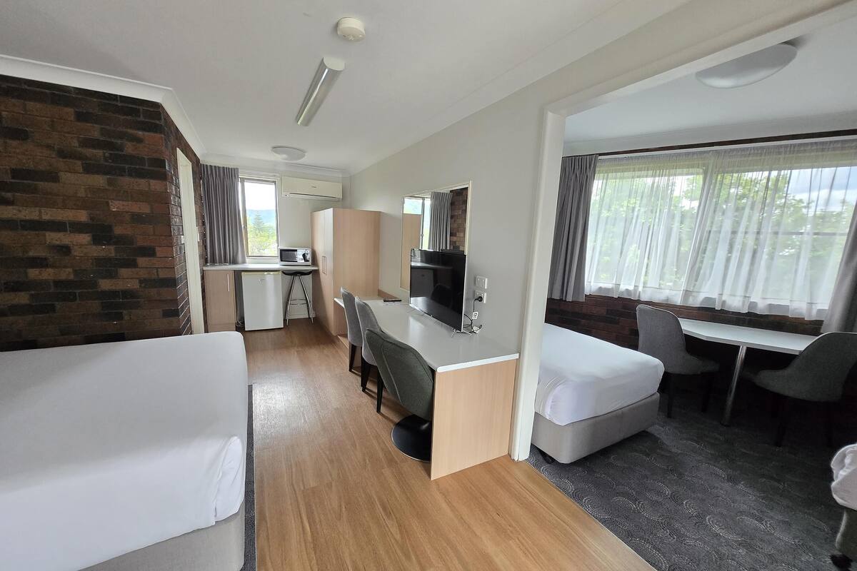 Quarto de hotel com uma cama, uma mesa, uma cadeira, uma TV e uma cozinha compacta.