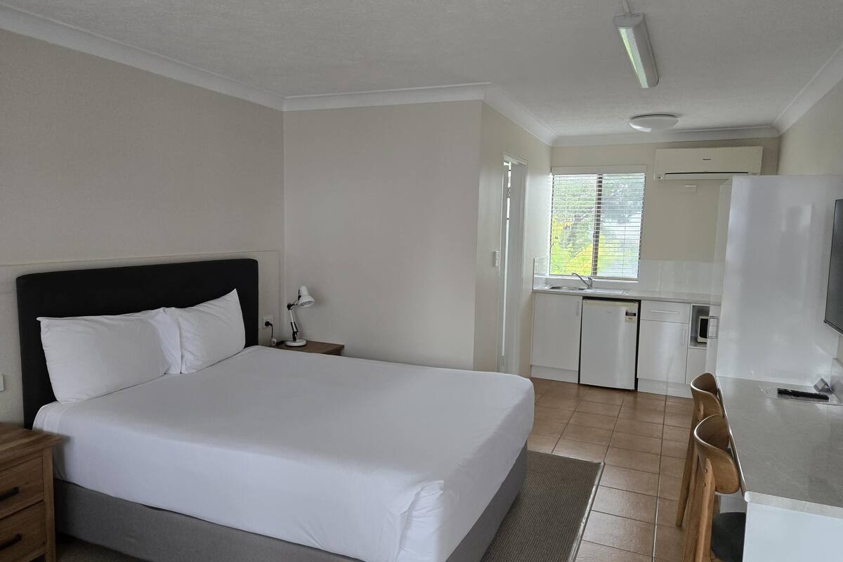 Quarto de hotel com uma cama grande, uma escrivaninha com cadeira, uma pequena cozinha e uma janela com persianas.