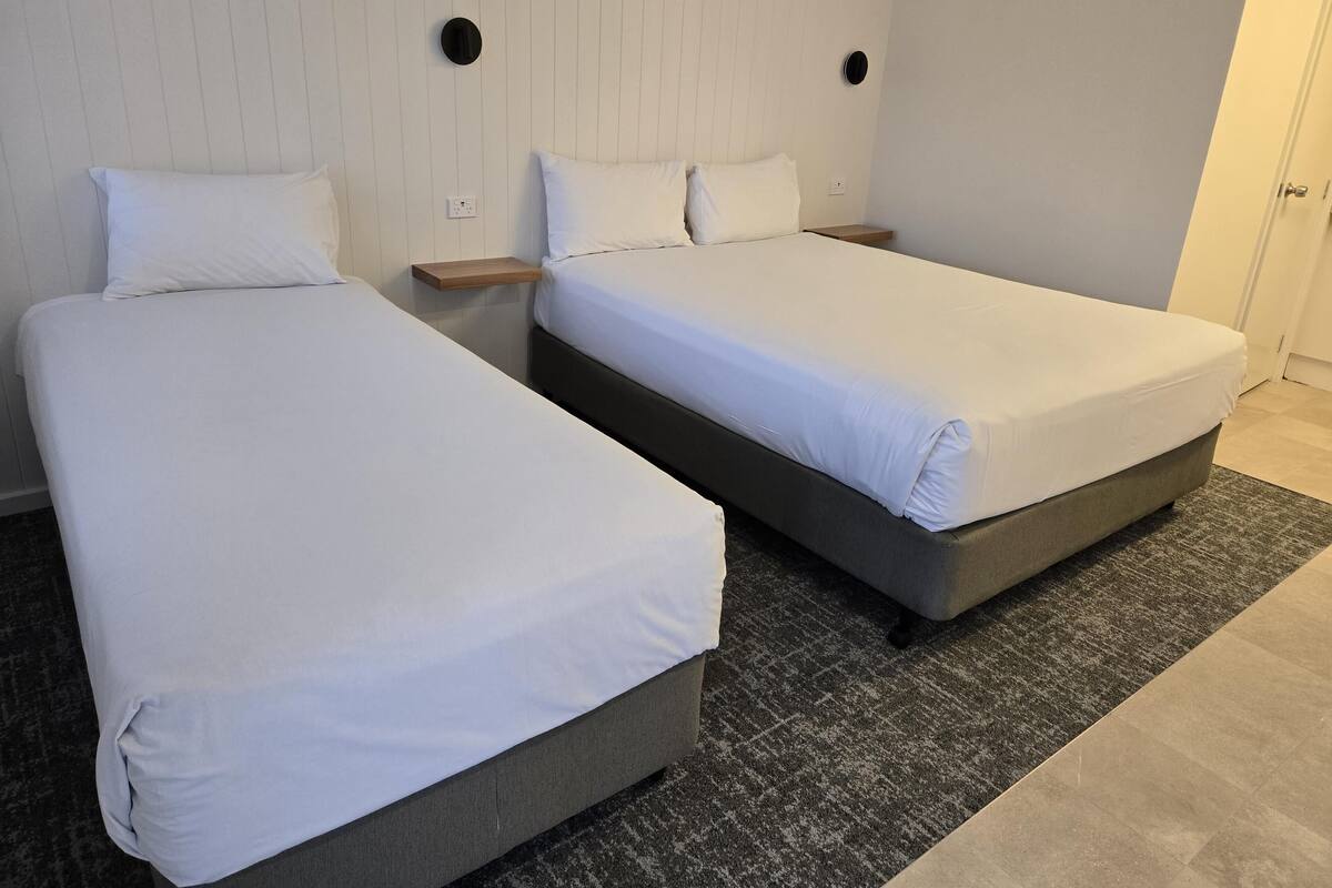 Quarto de hotel com duas camas de solteiro, uma pequena mesa de cabeceira de madeira, uma luminária fixada na parede e uma porta.