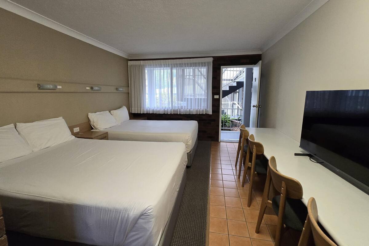 Quarto de hotel com duas camas, televisão, mesa de jantar com cadeiras e janela com cortinas.