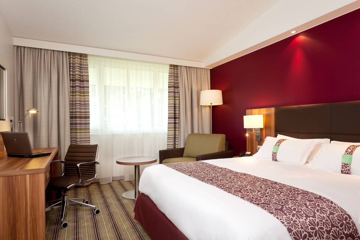 Quarto de hotel com uma cama grande, uma escrivaninha com um laptop, duas cadeiras, uma mesinha e um abajur.