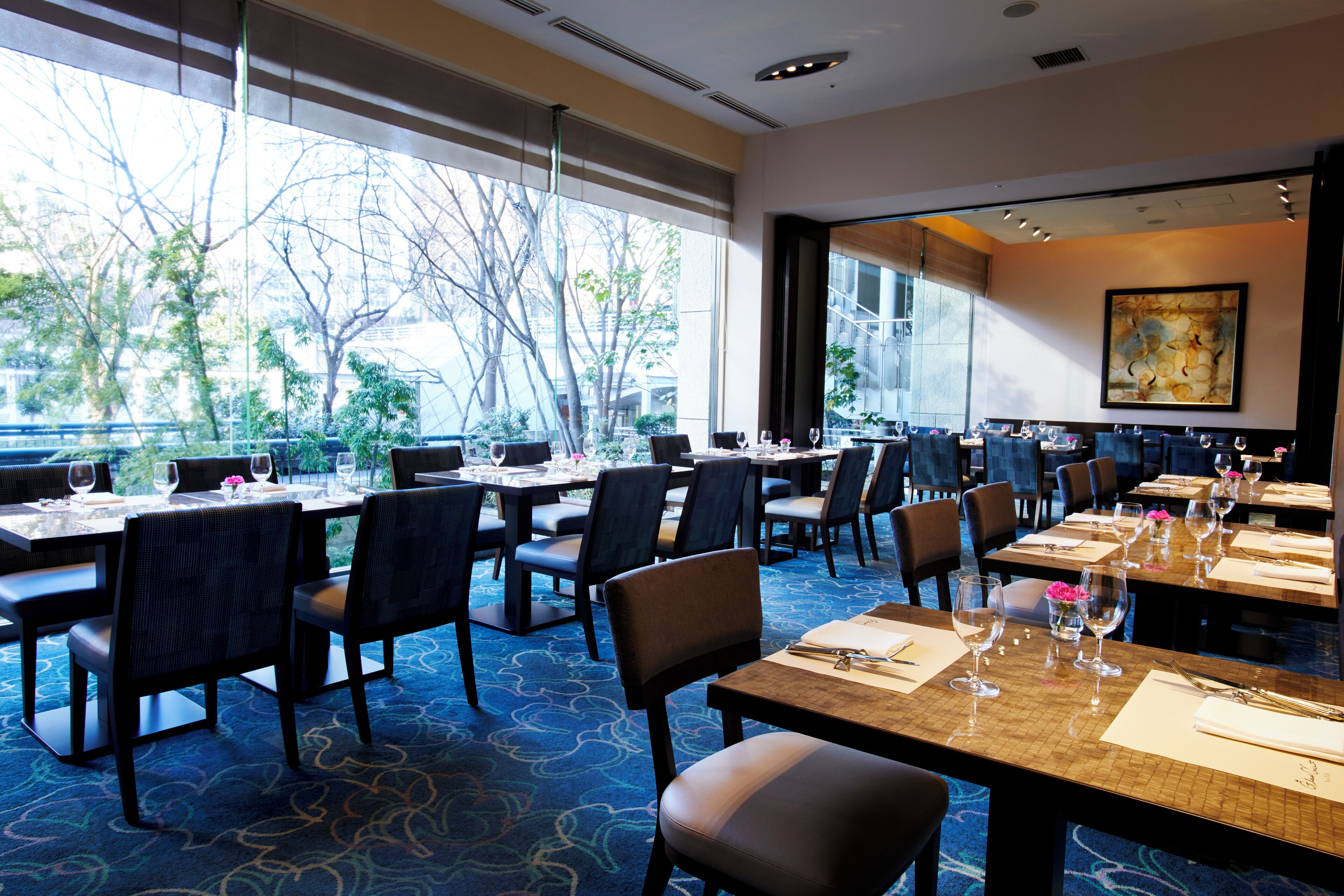 Restaurante com mesas postas para refeições, grandes janelas e vista para as árvores.
