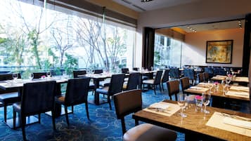 Restaurante com mesas postas para refeições, grandes janelas e vista para as árvores.