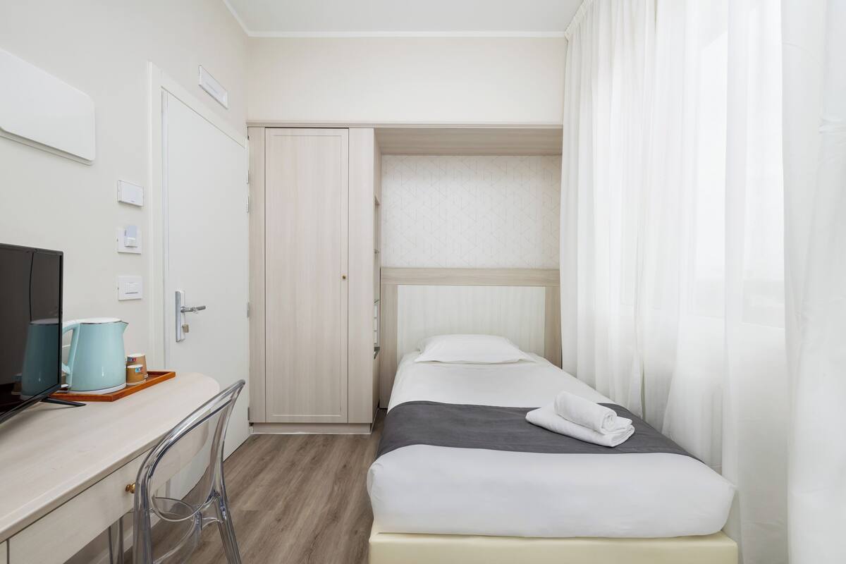 Quarto de hotel pequeno com uma cama, uma secretária com computador e um guarda-roupa.