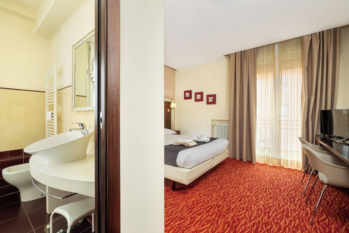 Quarto de hotel com banheiro, incluindo pia e espelho, uma cama sobre um tapete vermelho estampado e uma mesa de trabalho com cadeira.