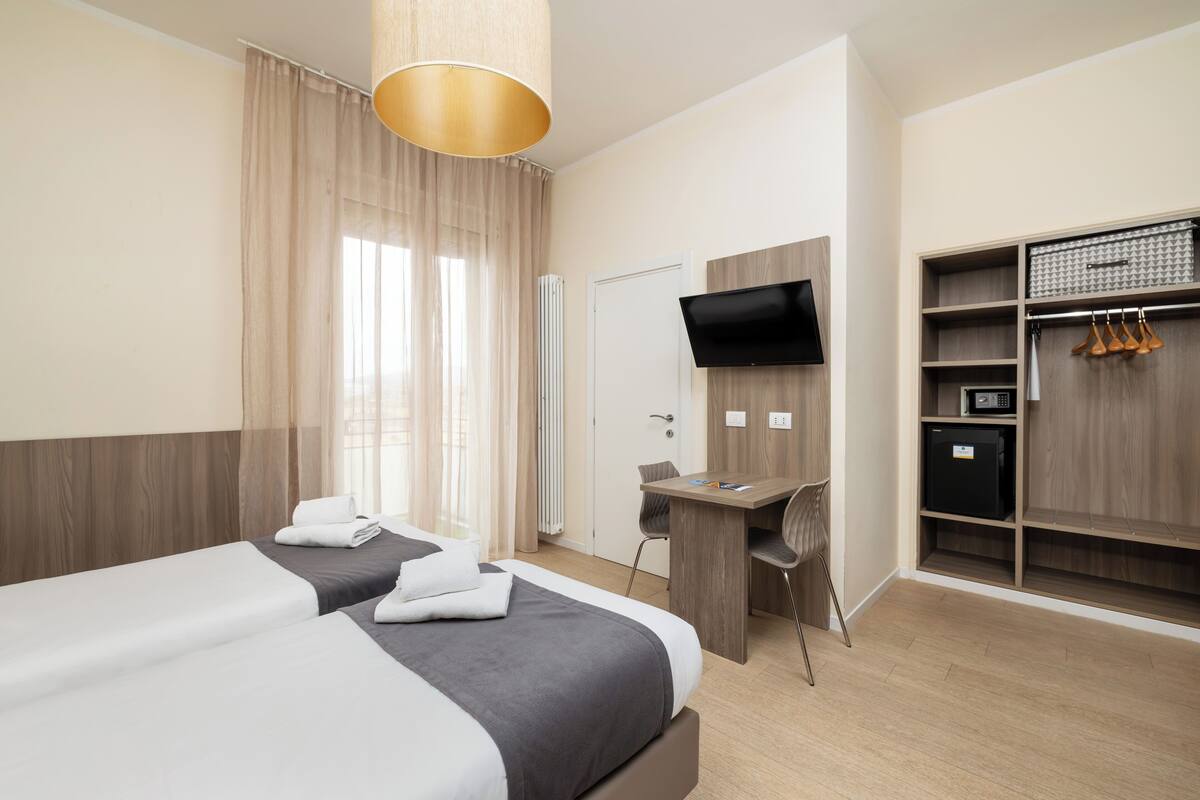 Quarto de hotel com duas camas, uma escrivaninha, uma cadeira, uma televisão, um guarda-roupa e um radiador.