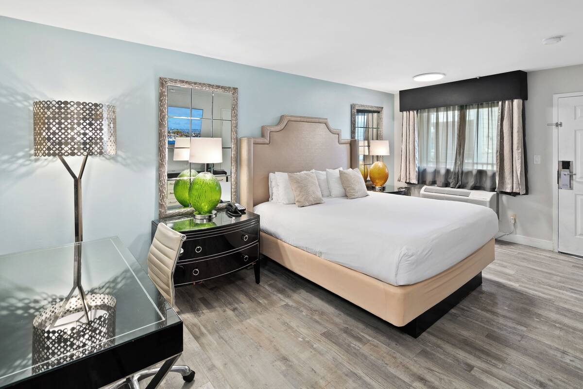 Quarto de hotel moderno com uma cama grande, uma escrivaninha de vidro, um espelho e uma janela com cortinas.