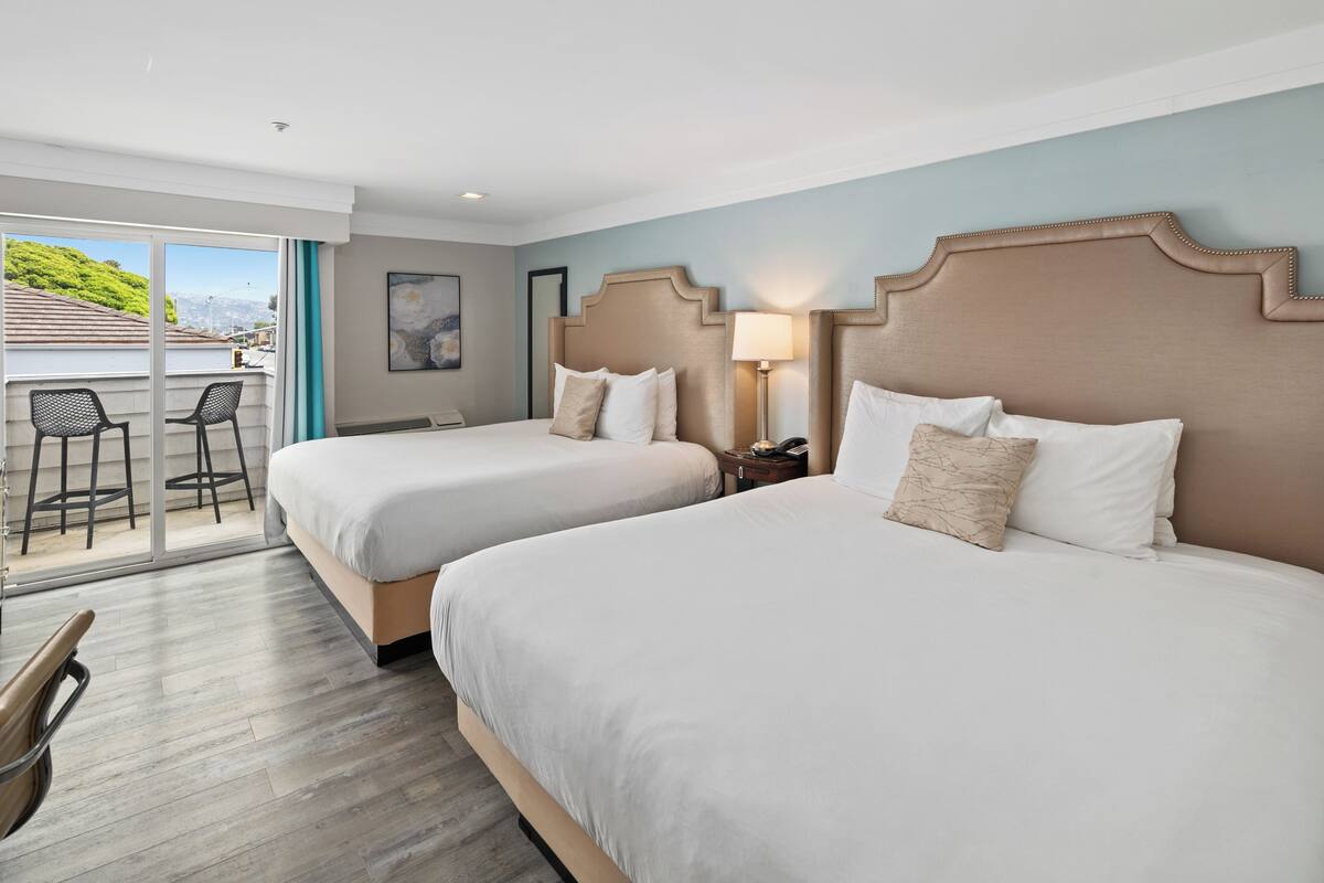 Quarto de hotel com duas camas, varanda com cadeiras e vista para a cidade.
