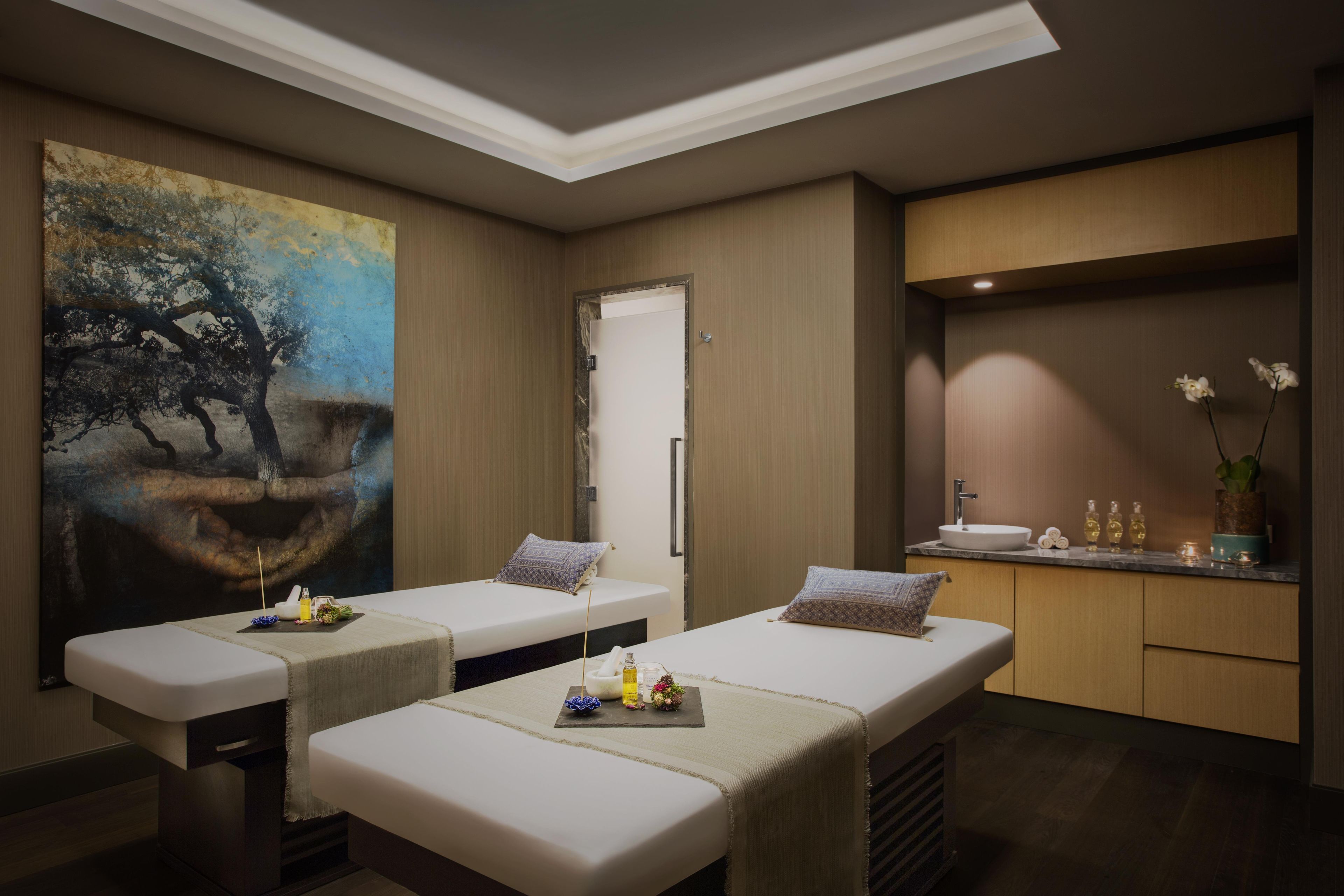 Sala de spa com duas macas de tratamento, uma pintura grande, uma pia e uma pequena planta.