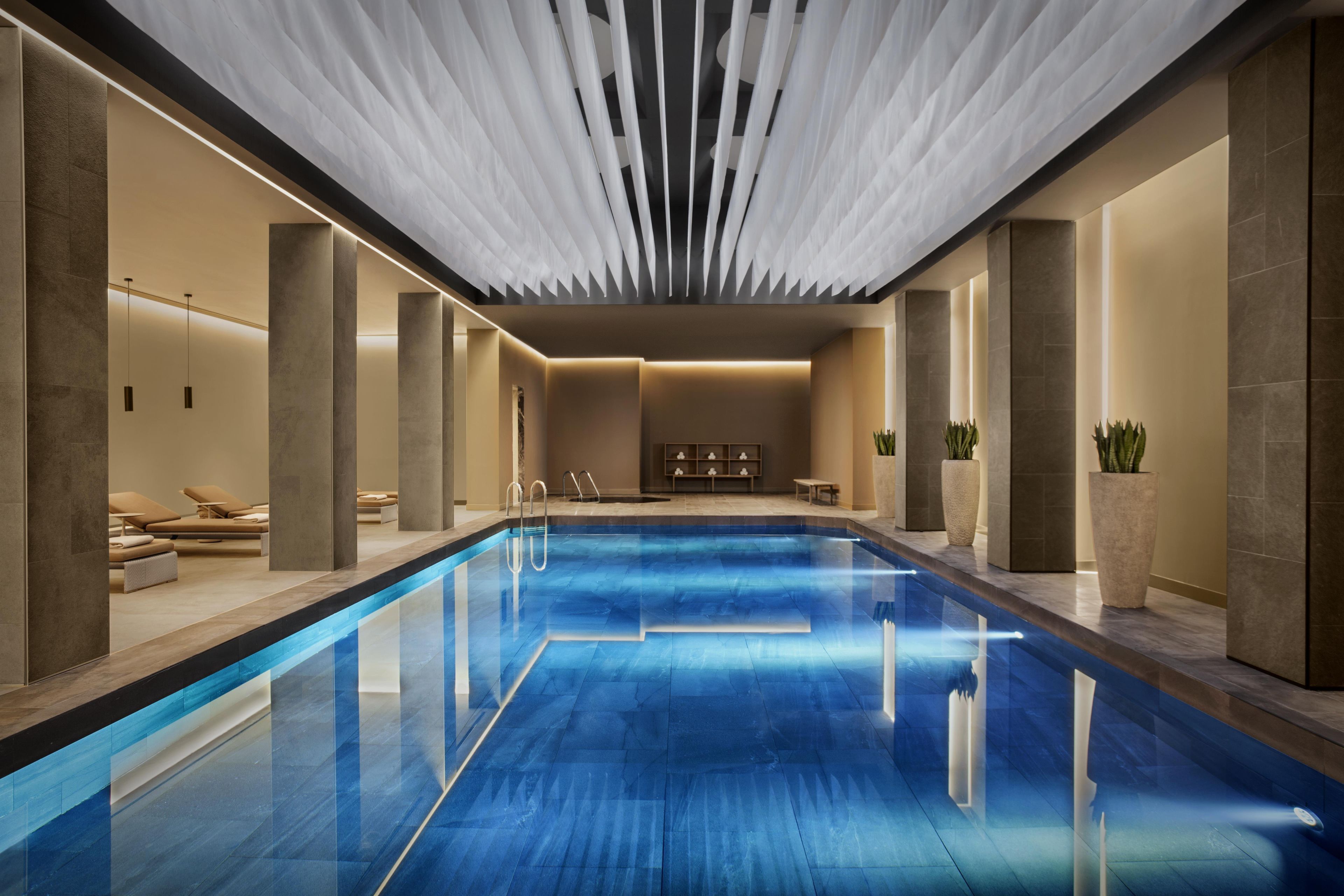 Piscina interior moderna com água azul cristalina, rodeada por paredes e colunas em tons de bege, e com luminária no teto.