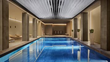 Piscina interior moderna com água azul cristalina, rodeada por paredes e colunas em tons de bege, e com luminária no teto.