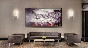 Um saguão com uma grande pintura de paisagem montanhosa, um sofá cinzento, duas poltronas e uma mesa de centro com um vaso de flores.