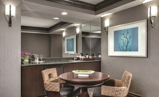 Quarto de hotel moderno com mesa de jantar redonda, duas poltronas estofadas, área de bar com pia e um espelho grande que reflete o quarto.