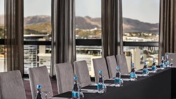 Mesa de conferência com cadeiras, garrafas de água e copos, com montanhas e paisagem urbana ao fundo.