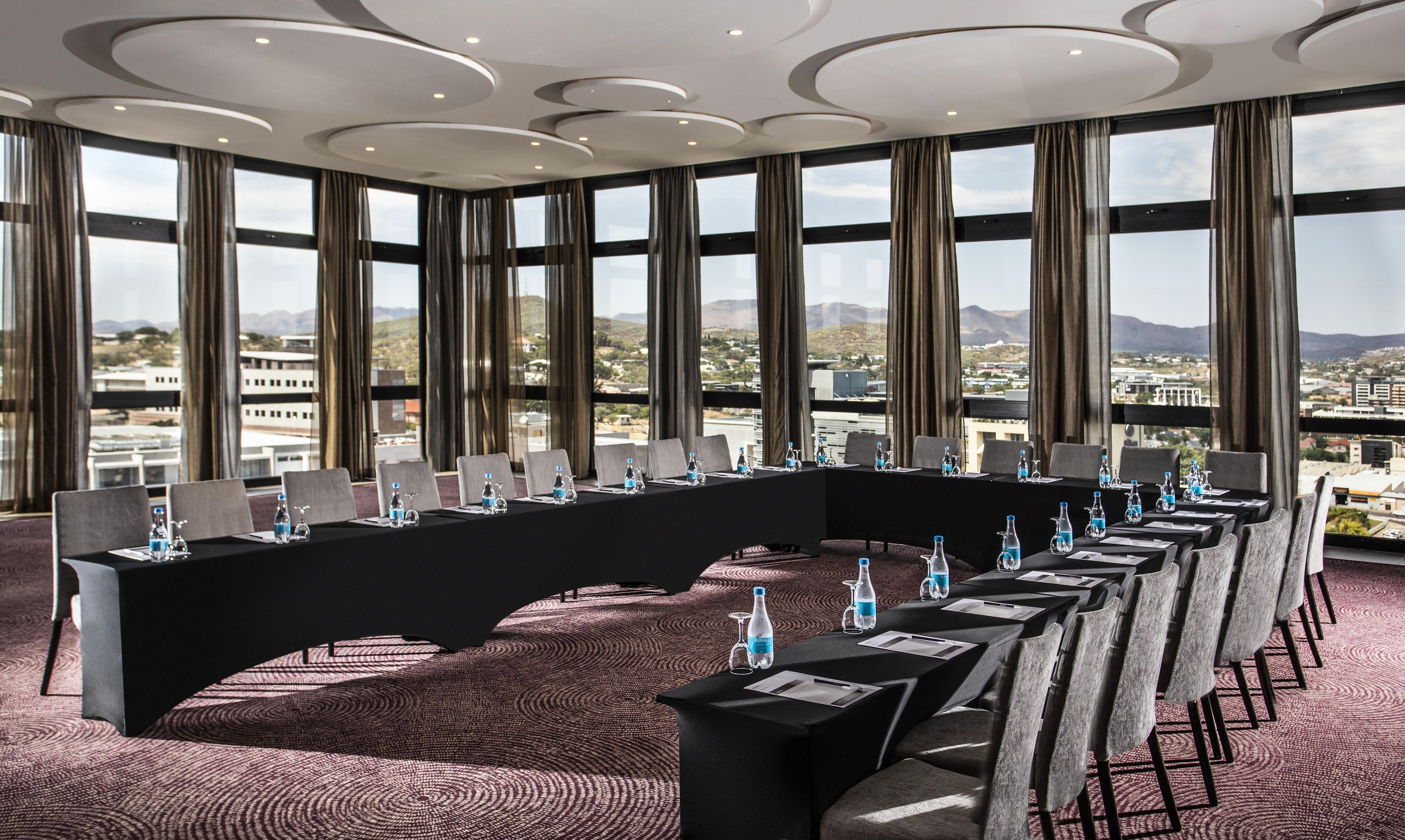 Sala de conferências com uma mesa comprida, cadeiras e garrafas de água, e vista para a cidade através de grandes janelas.