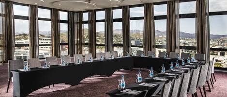 Sala de conferências com uma mesa comprida, cadeiras e garrafas de água, e vista para a cidade através de grandes janelas.
