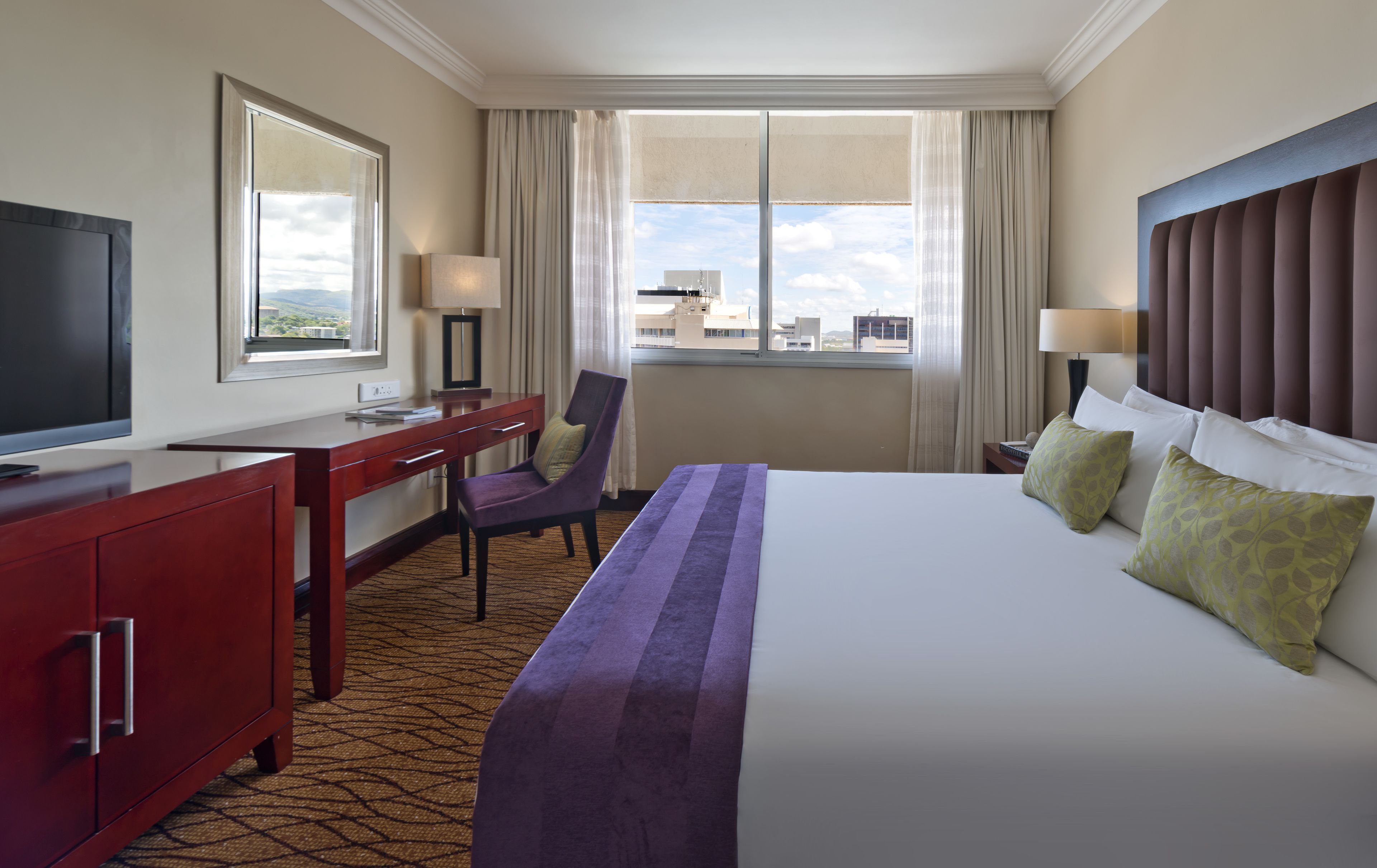 Quarto de hotel com cama, escrivaninha, cadeira, televisão e janela com vista para a cidade.