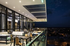 Terraço ao ar livre com assentos e mesas, com vista para a paisagem urbana à noite.