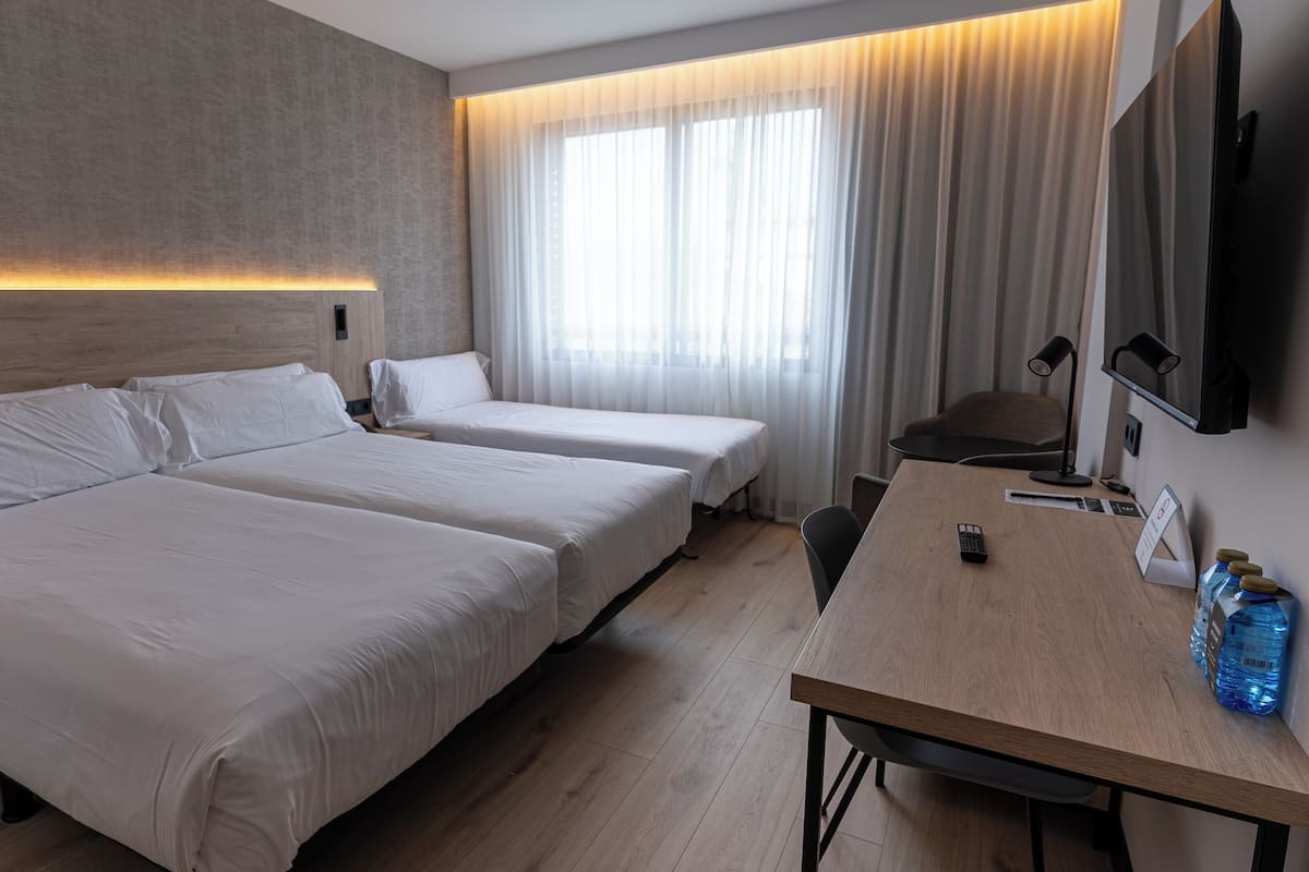 Quarto de hotel com duas camas, uma escrivaninha, uma cadeira, televisão de tela plana e janela com cortinas.