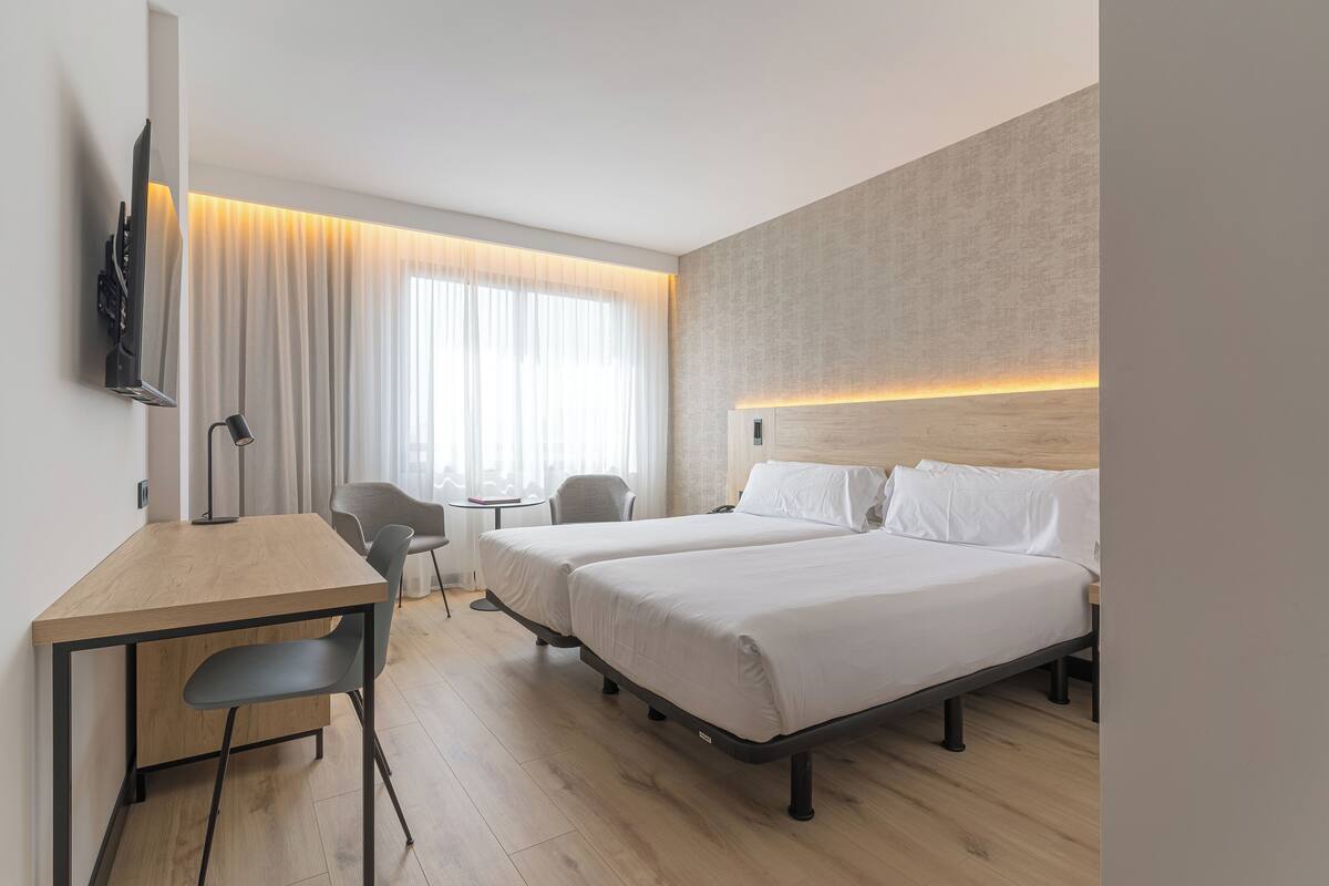Quarto de hotel com escrivaninha de madeira, duas camas de solteiro, televisão fixada na parede e uma janela grande com cortinas.