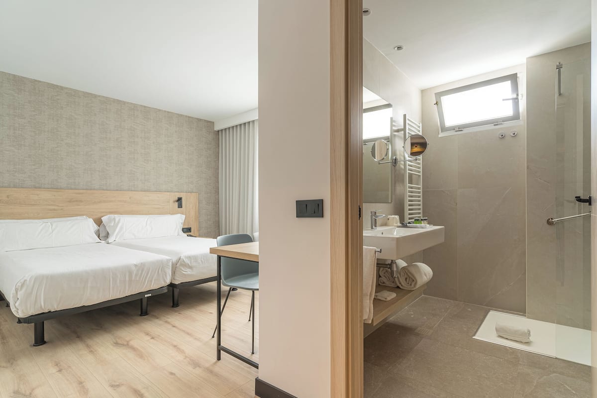 Quarto de hotel com duas camas, uma escrivaninha, chuveiro e janela.