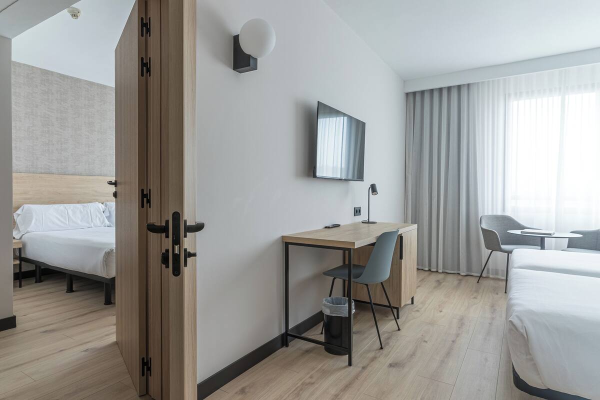 Quarto de hotel com escrivaninha de madeira, duas camas de solteiro, televisão e uma janela grande com cortinas.