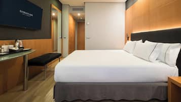 Quarto de hotel com cama, escrivaninha com cafeteira e televisão fixada na parede.