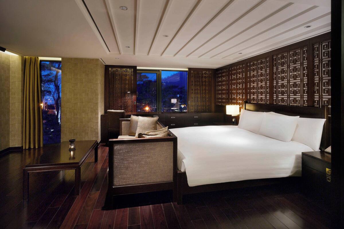 Quarto de hotel moderno com uma cama grande, mesa de cabeceira e vista para a cidade à noite.