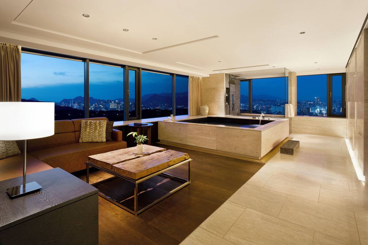 Quarto de hotel moderno com uma banheira grande, um sofá, uma mesa de centro e vista para a cidade ao entardecer.
