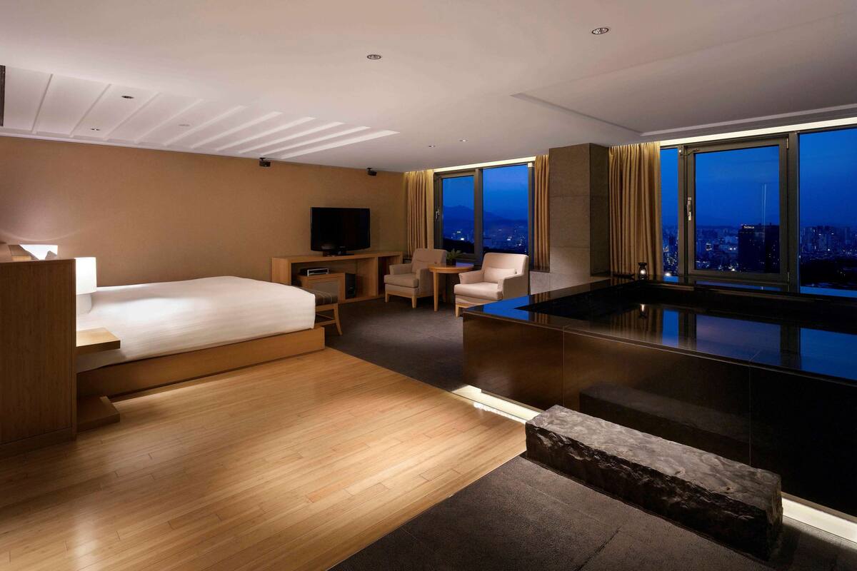Quarto de hotel moderno com cama, área de estar, TV e uma grande janela com vista para a cidade.
