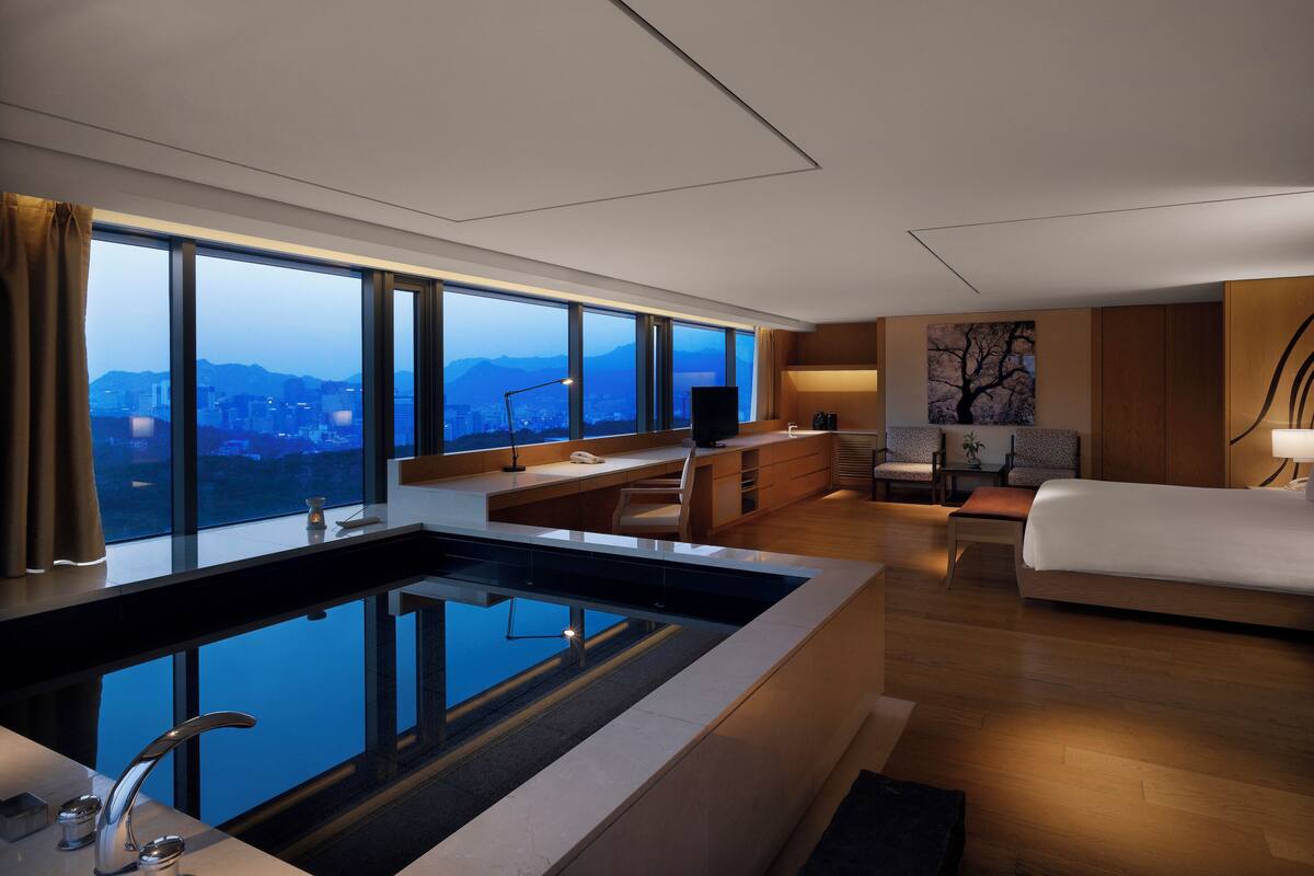Quarto de hotel moderno com uma janela grande, uma banheira, um sofá e uma mesa de trabalho com televisão.