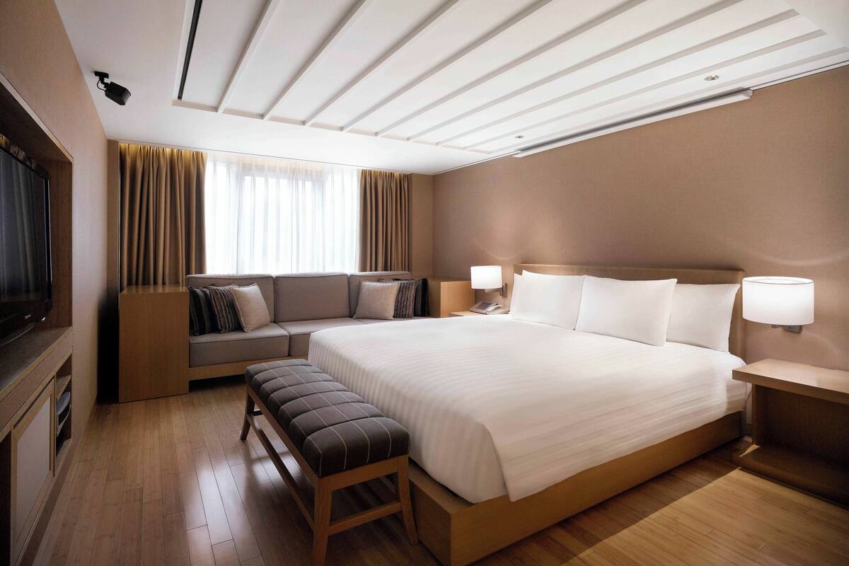 Quarto de hotel com uma cama grande, um sofá, um banco, uma TV e uma escrivaninha com abajur.