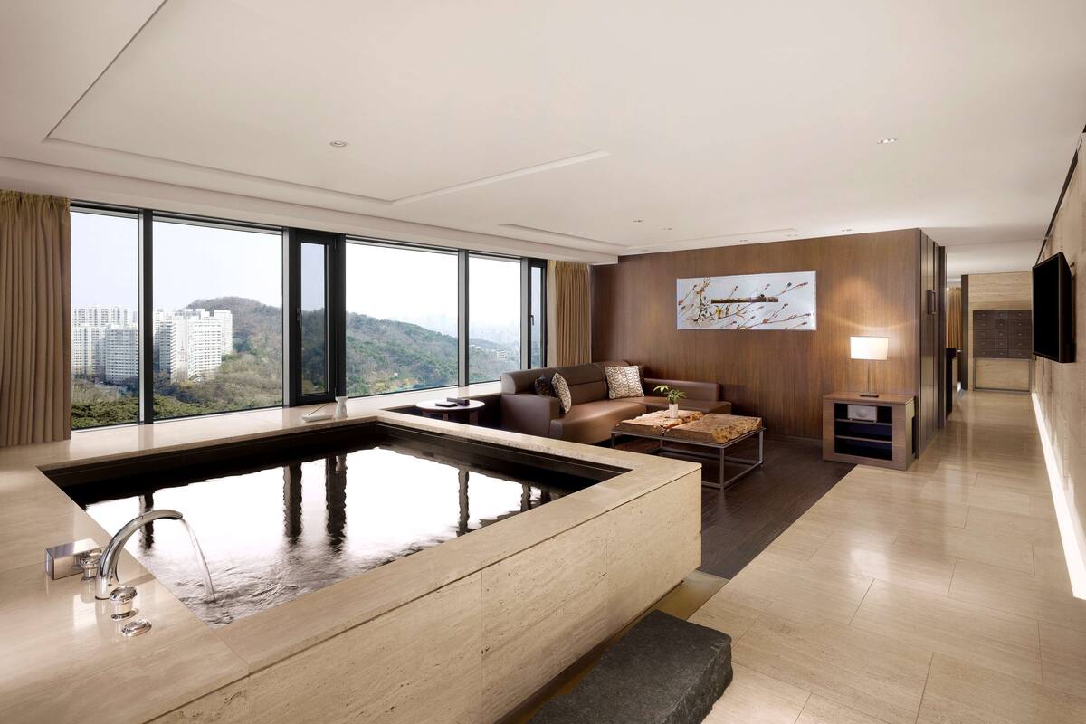 Quarto de hotel moderno com uma janela ampla, hidromassagem integrada, sofá e televisão de tela plana.