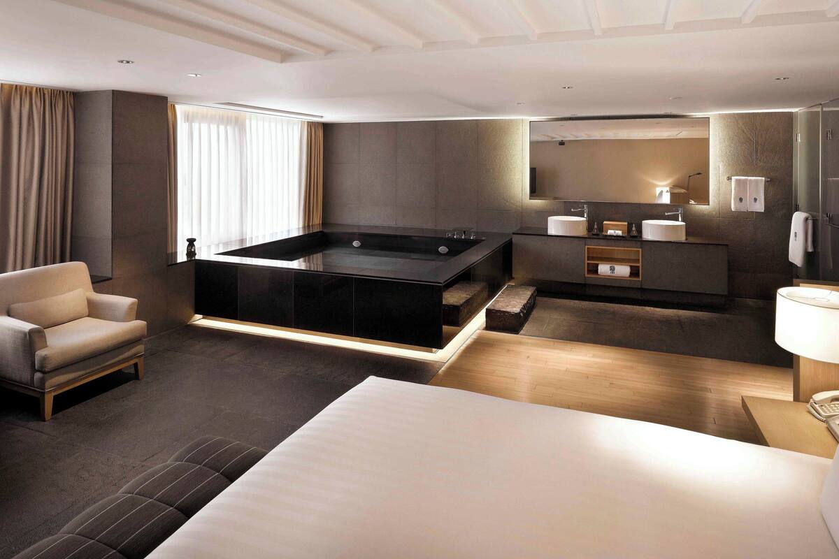Quarto de hotel moderno com uma cama grande, um sofá, uma escrivaninha, banheiro com banheira e um espelho amplo.