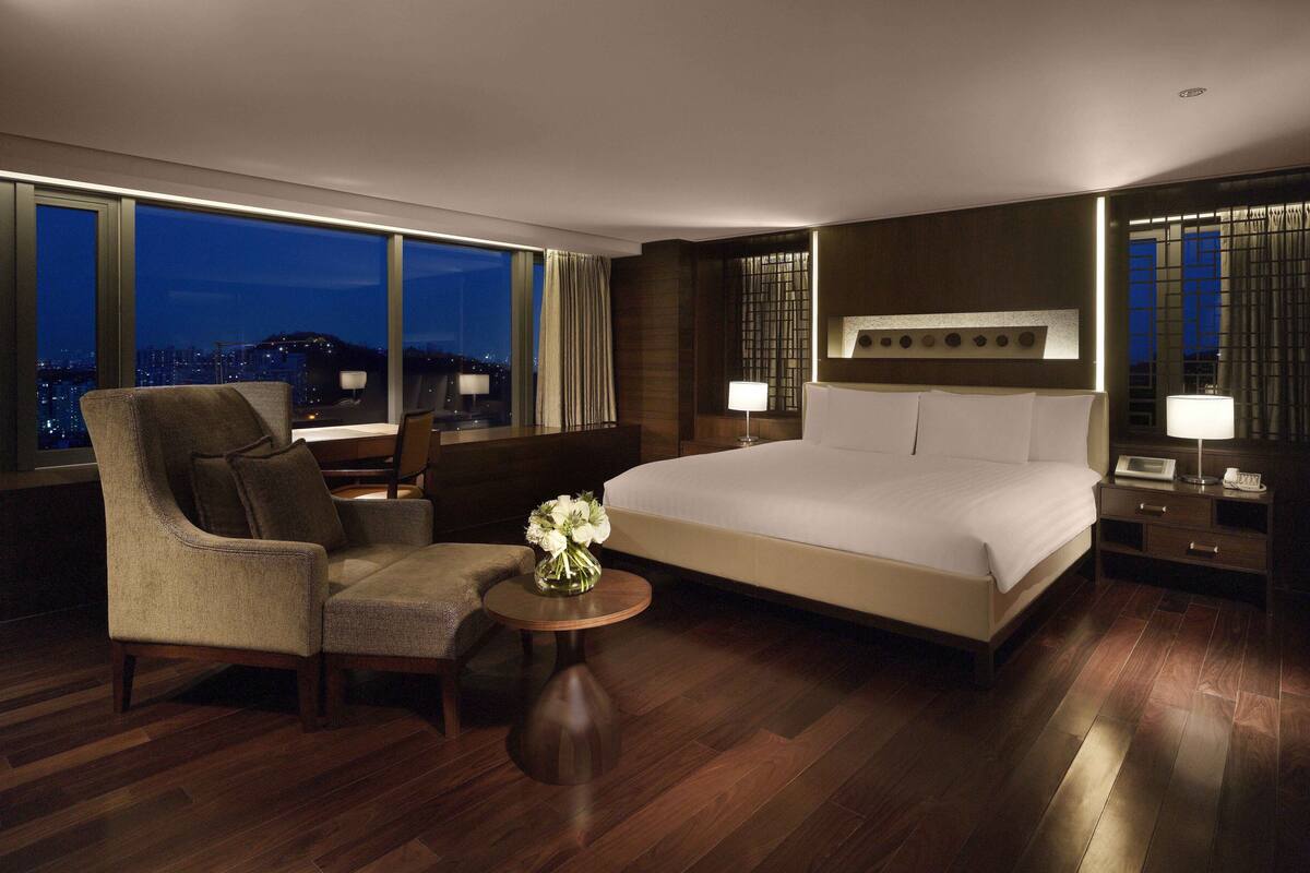 Quarto de hotel com uma cama grande, área de estar com uma cadeira e uma mesinha, e vista para a cidade à noite.