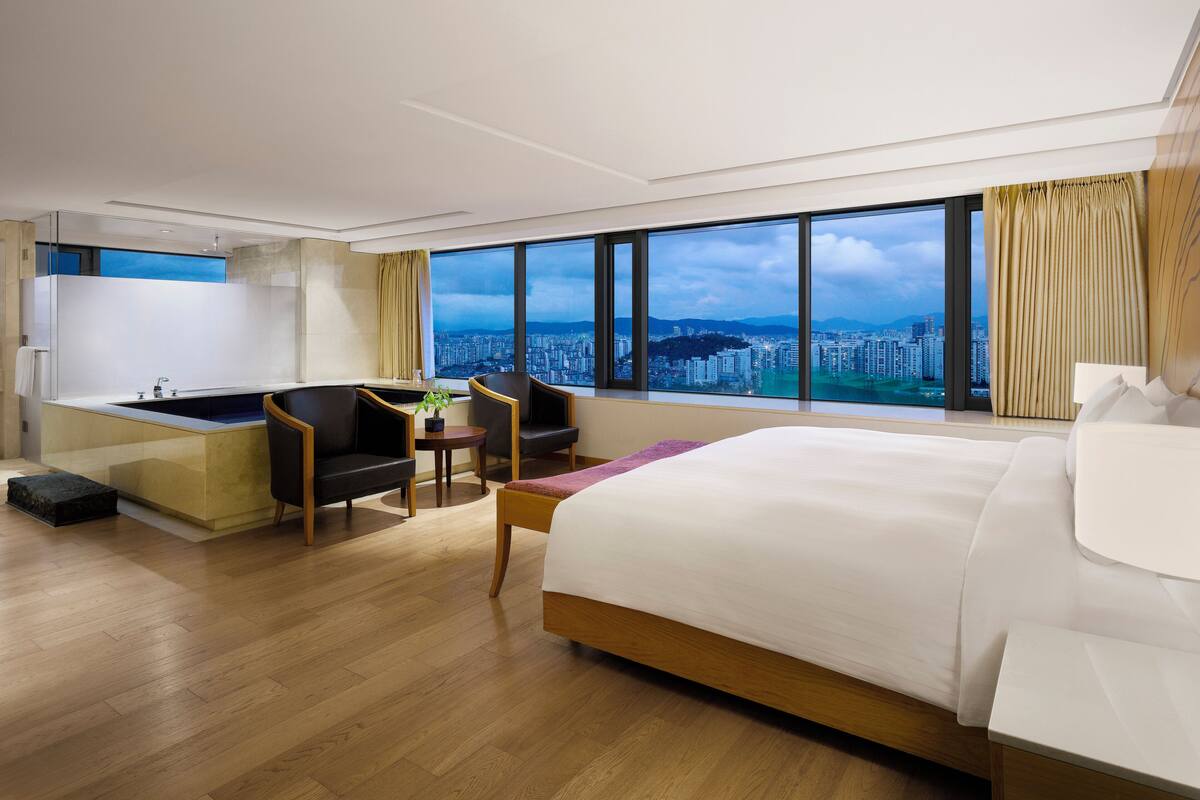 Quarto de hotel com cama, escrivaninha com cadeira, pequena mesa com planta e uma grande janela com vista para a cidade.