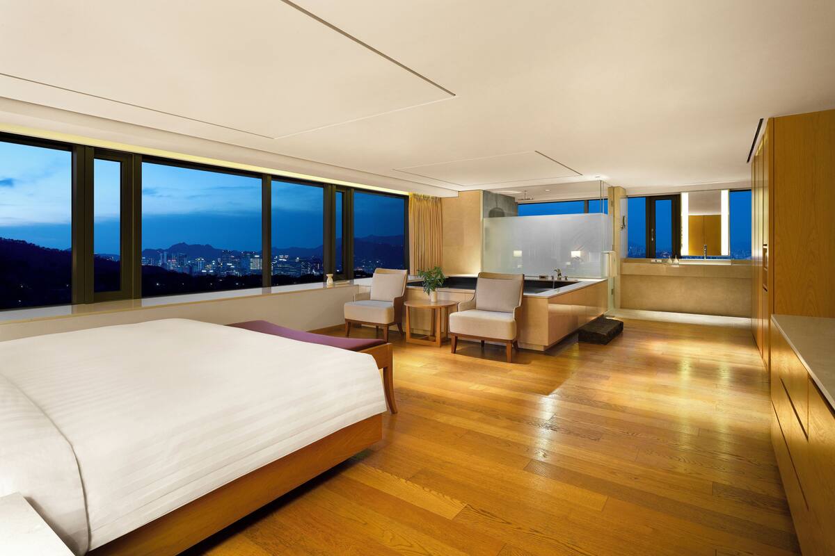 Quarto de hotel moderno com uma cama grande, uma escrivaninha, uma cadeira e vista para a paisagem urbana.