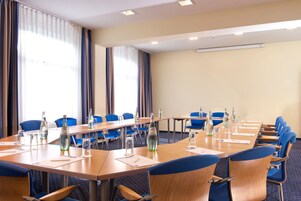 Uma sala de conferências com mesas compridas, cadeiras azuis e garrafas de água.