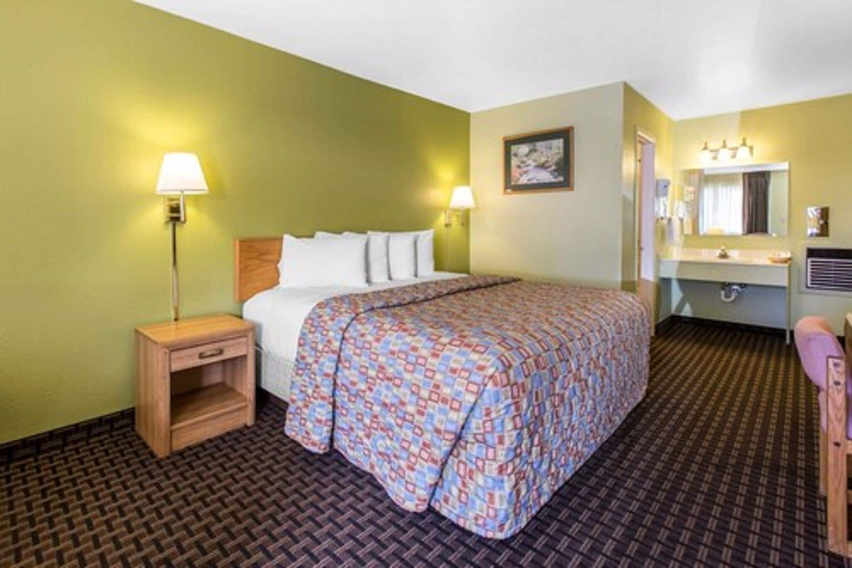 Quarto de hotel com cama, luminárias de cabeceira, mesa de cabeceira de madeira, banheiro com pia e espelho, e tapete estampado.