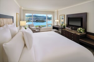 Quarto de hotel com cama grande, televisão, escrivaninha e vista para um lago e montanhas.