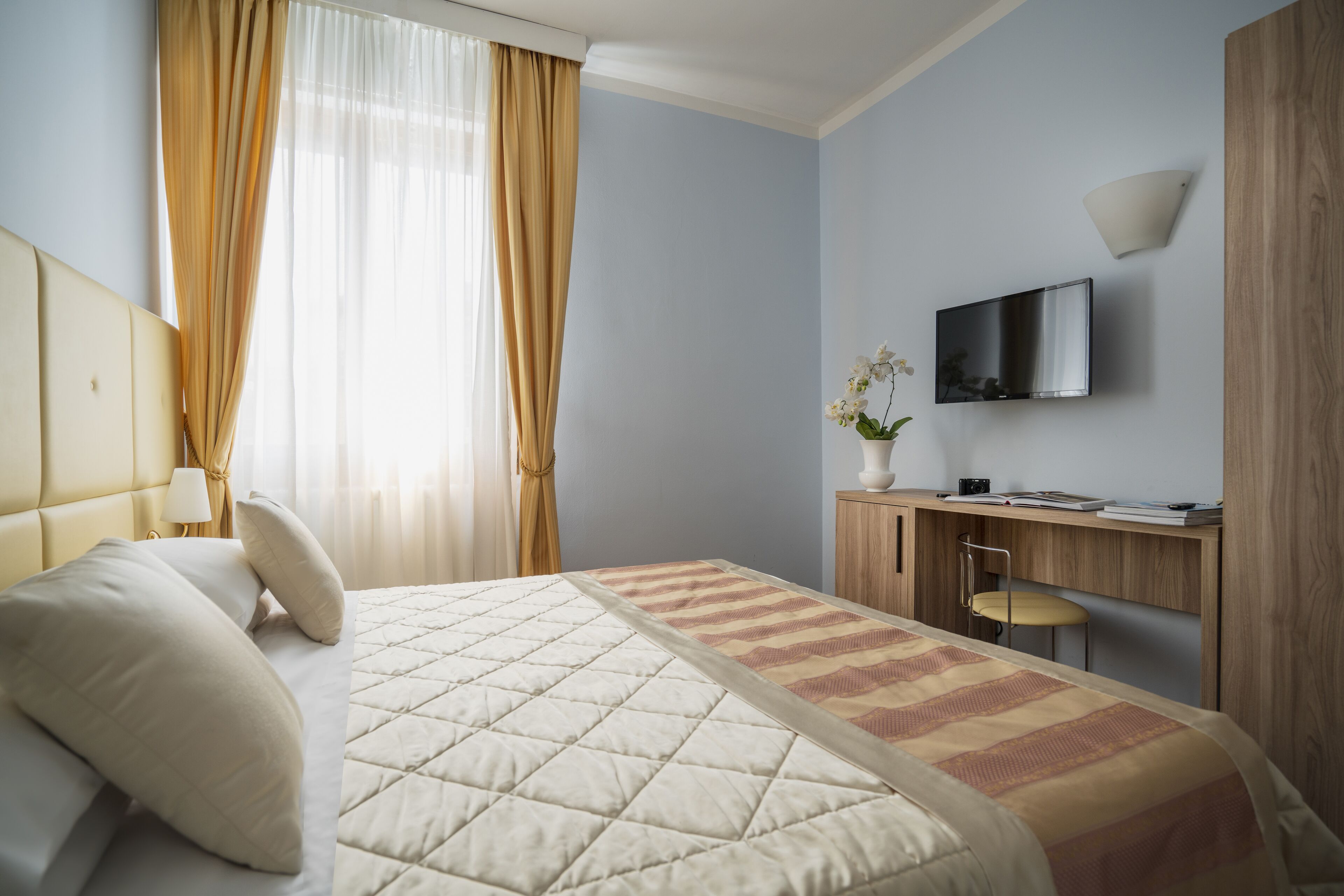 Quarto de hotel com cama, escrivaninha, televisão e janela com cortinas.